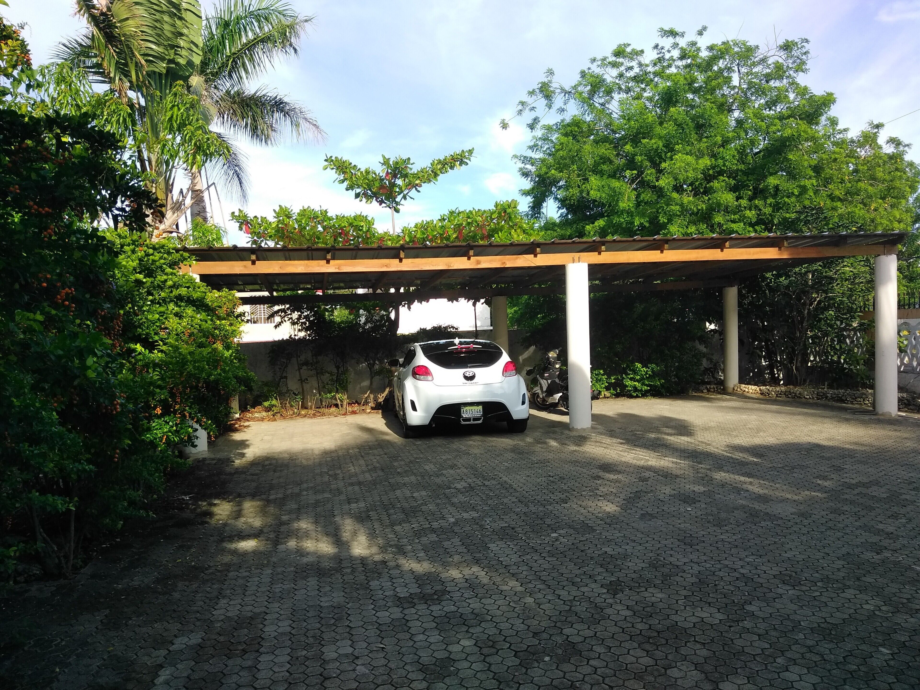 Área de estacionamento coberta com um carro branco estacionado sob uma pérgula de madeira.