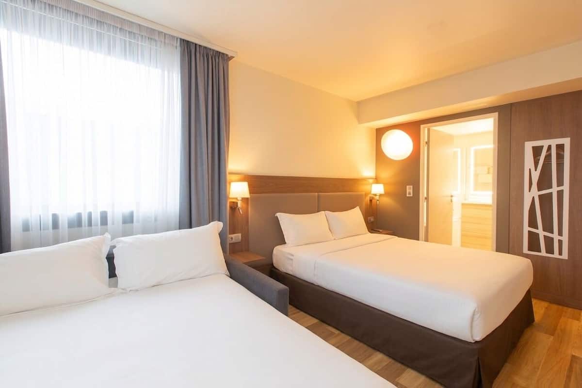 Quarto de hotel com duas camas, uma janela grande com cortinas, luminárias de cabeceira e uma porta com design geométrico.
