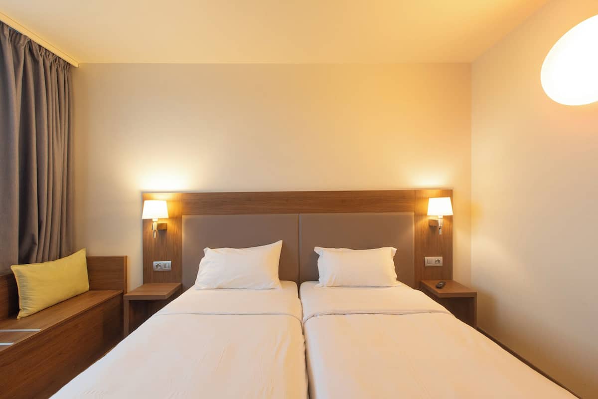 Quarto de hotel com duas camas, cabeceiras de madeira, luminárias de cabeceira, mesa de cabeceira com controle remoto e um travesseiro amarelo.