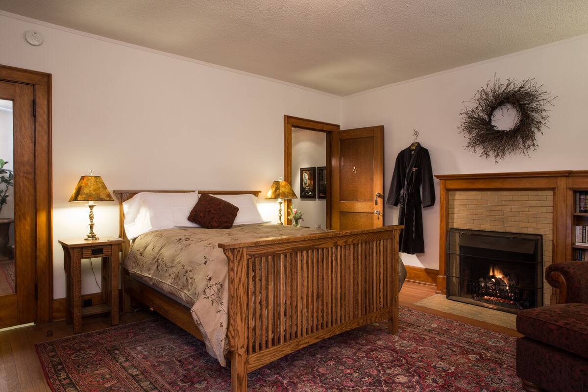 Quarto com cama de madeira, duas luminárias de cabeceira, lareira, espelho e porta-casacos.