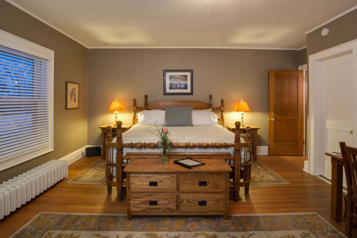 Quarto com cama de madeira, abajures, mesa de cabeceira, um quadro na parede e radiador sob a janela.