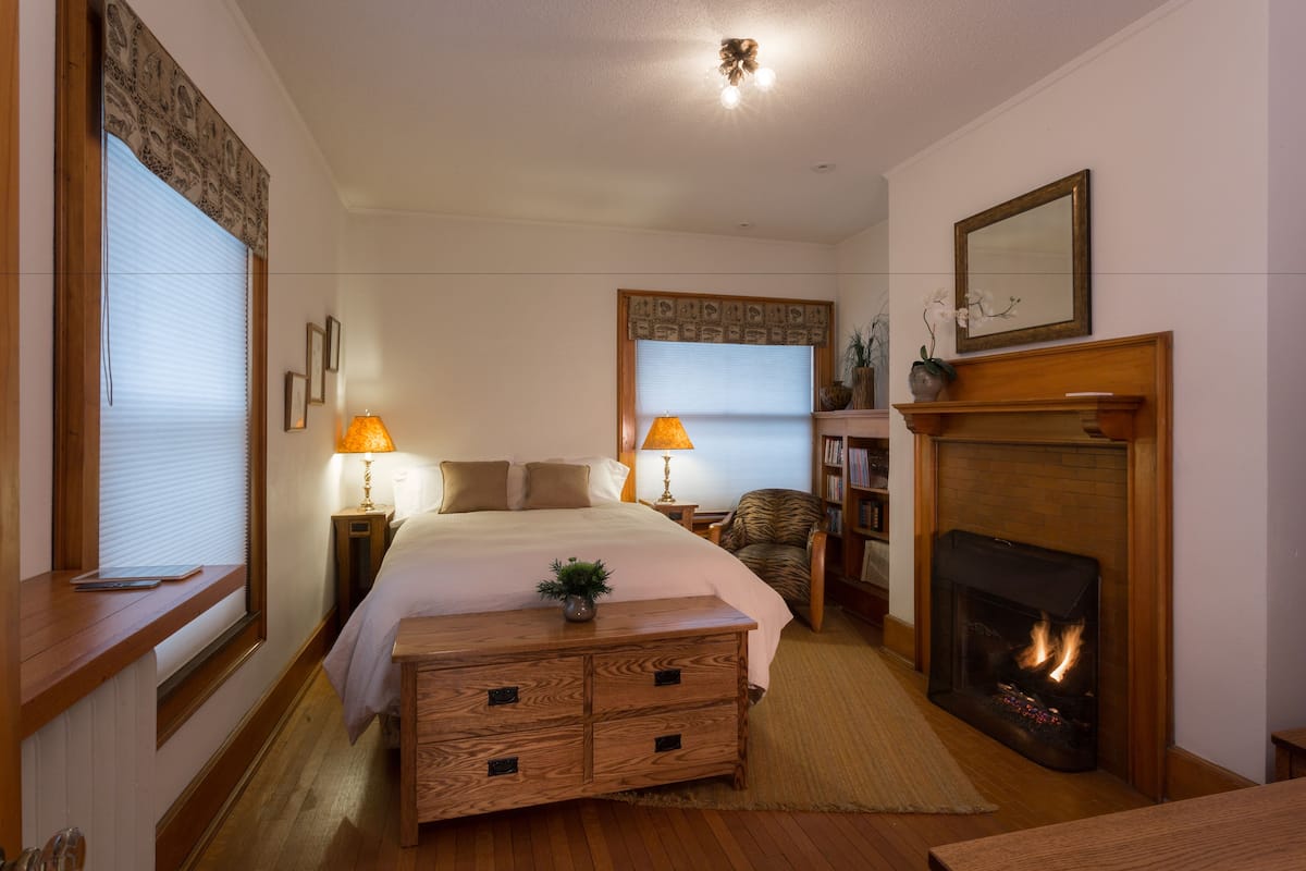 Quarto com cama, abajures, cómoda de madeira, lareira e estante de livros.