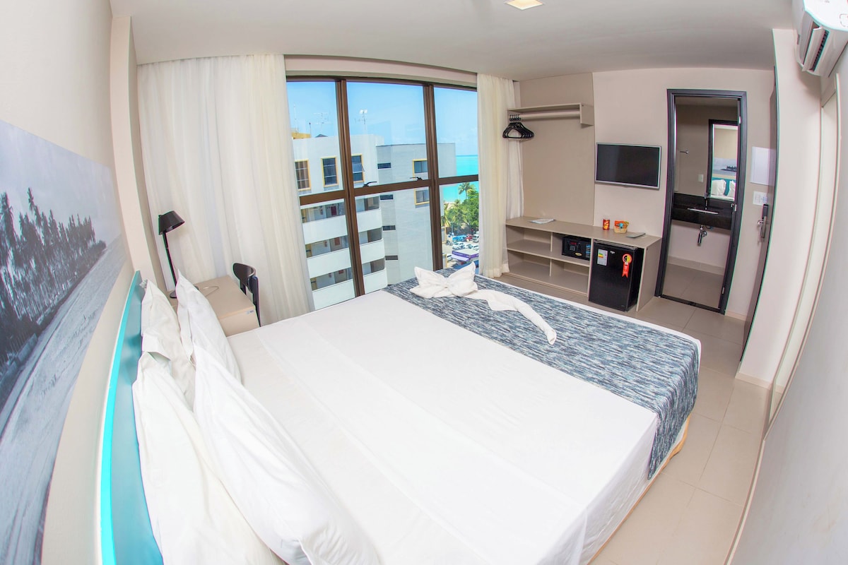 Quarto de hotel com uma cama grande, uma mesa, uma TV e vista da cidade através de grandes janelas.