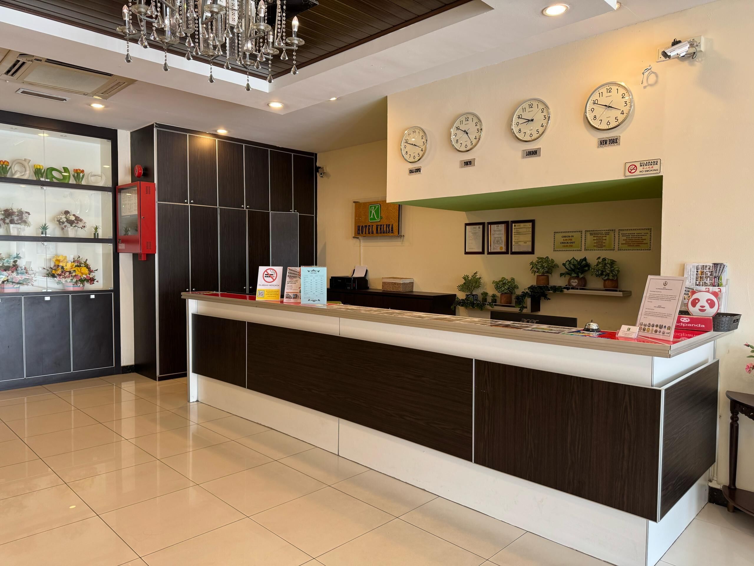 Uma área de recepção de hotel com design moderno, apresentando um balcão de atendimento, relógios de parede exibindo diferentes fusos horários e um candelabro.