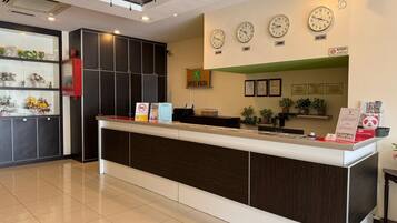 Uma área de recepção de hotel com design moderno, apresentando um balcão de atendimento, relógios de parede exibindo diferentes fusos horários e um candelabro.