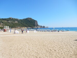 Uma praia com espreguiçadeiras e guarda-sóis, uma rede de voleibol e uma colina rochosa ao fundo.
