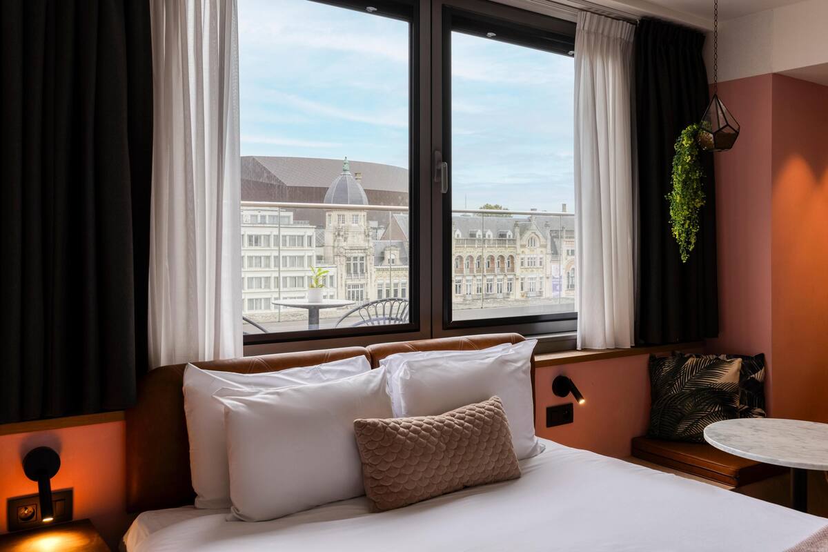 Quarto de hotel com uma cama, uma mesinha, uma planta pendente e vista para edifícios históricos.