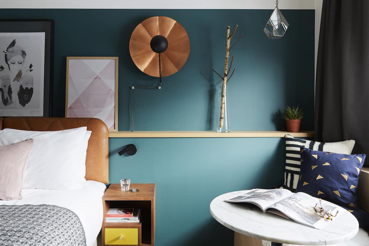 Quarto bem arrumado com cama, mesa de cabeceira e uma pequena mesa de leitura com um livro e óculos.