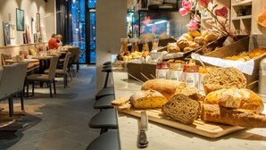 Vitrine de padaria com diversos tipos de pão e doces, uma faca em tábua de corte e uma pessoa ao fundo.