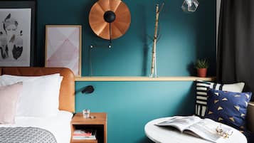 Quarto de hotel bem arrumado com cama, mesa de cabeceira, área de leitura e luminária na parede.