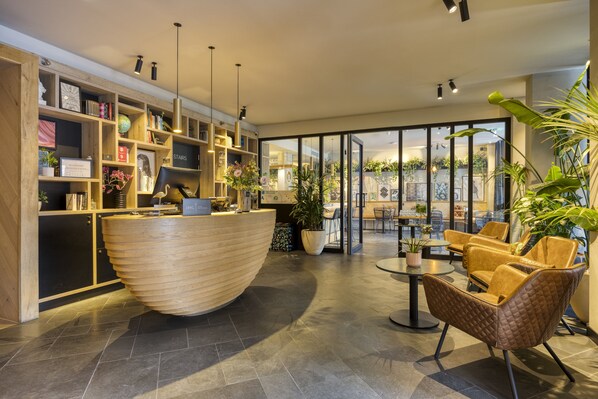 Uma área de recepção moderna com balcão de madeira, uma grande planta em vaso e uma sala de estar com poltronas de couro.