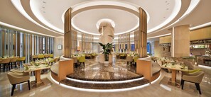 Um saguão de hotel moderno com uma escadaria central, área de refeições e cozinha ao fundo.