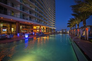 Uma área de piscina de hotel com uma piscina grande, espreguiçadeiras e um bar com mesas ao ar livre.