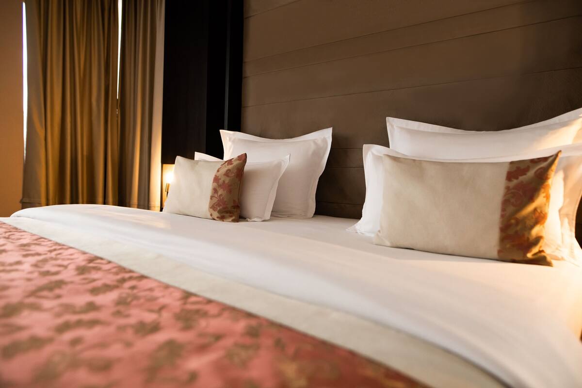 Cama bem arrumada com lençóis brancos, almofadas decorativas, colcha estampada e cabeceira de madeira.