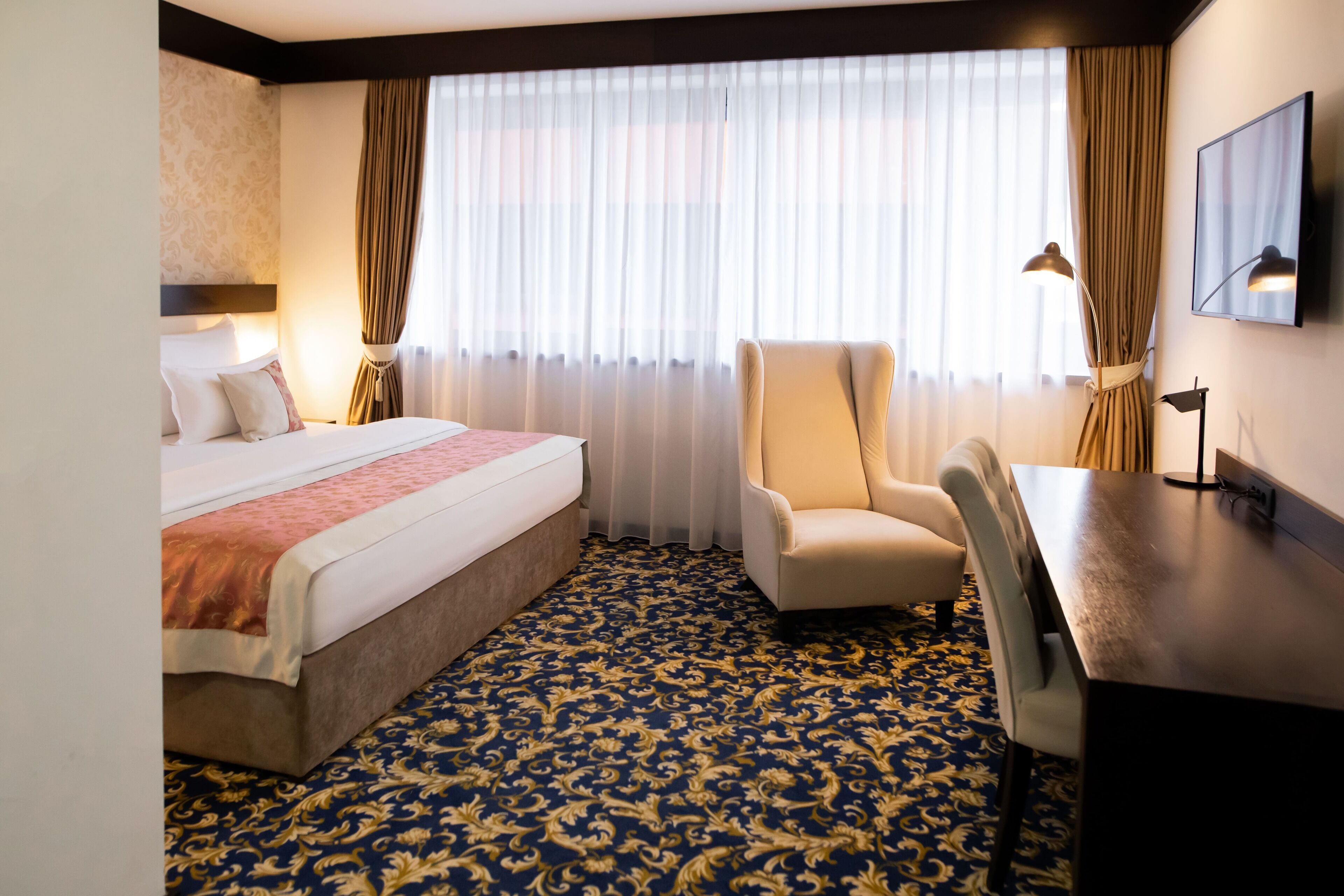 Quarto de hotel com uma cama grande, uma escrivaninha, uma cadeira e uma televisão na parede.