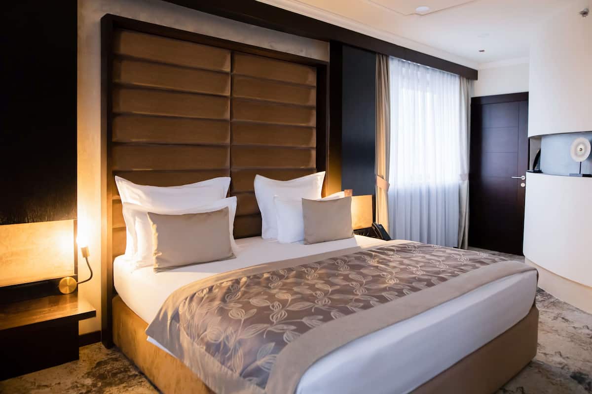 Quarto de hotel com uma cama grande, cabeceira de madeira, mesa de cabeceira com abajur e porta com moldura escura.