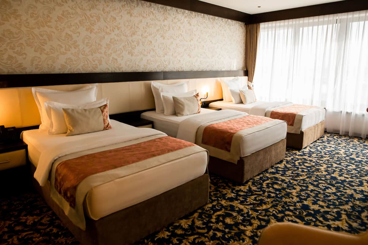 Quarto de hotel com três camas, um tapete estampado e uma janela grande com cortinas.