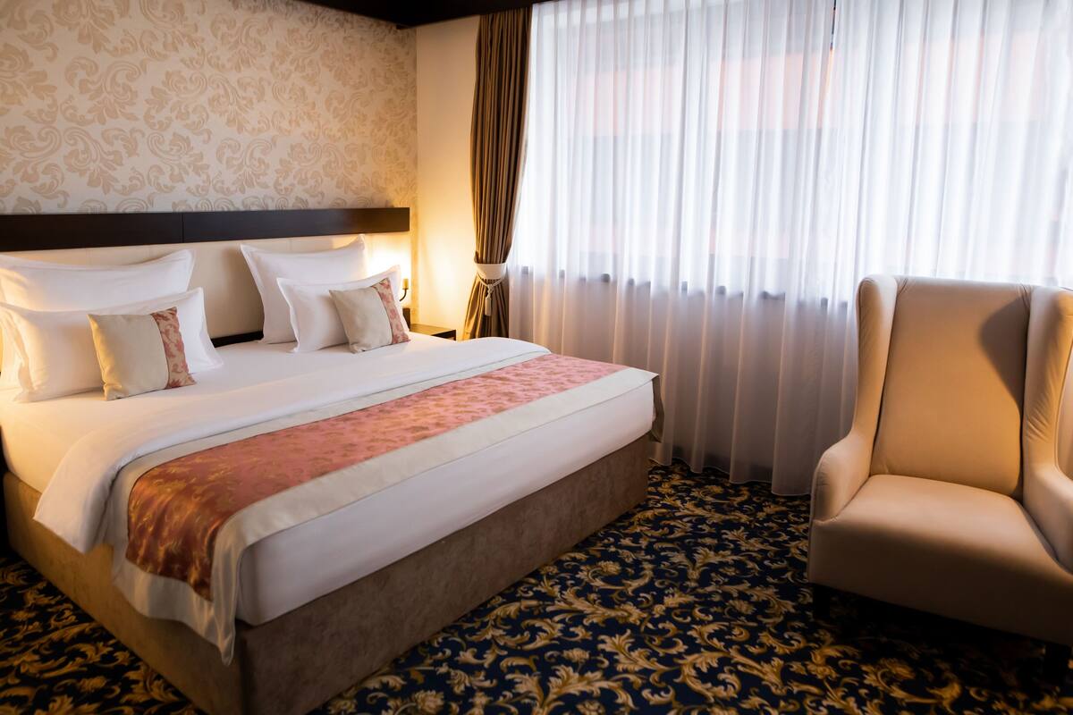 Quarto de hotel com uma cama grande, cabeceira, uma cadeira e um tapete estampado.