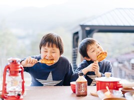 Duas crianças comendo à mesa, com um candeeiro, uma garrafa e uma panela vermelha.
