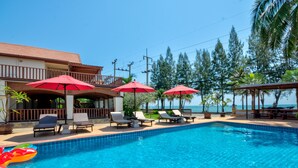 Piscina com espreguiçadeiras e guarda-sóis vermelhos, rodeada por um edifício com varanda com vista para o mar.