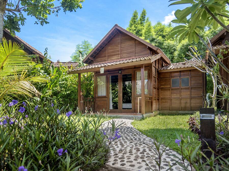 Uma casa de madeira com telhado de telhas, rodeada por vegetação exuberante e um caminho com padrão que leva à entrada.