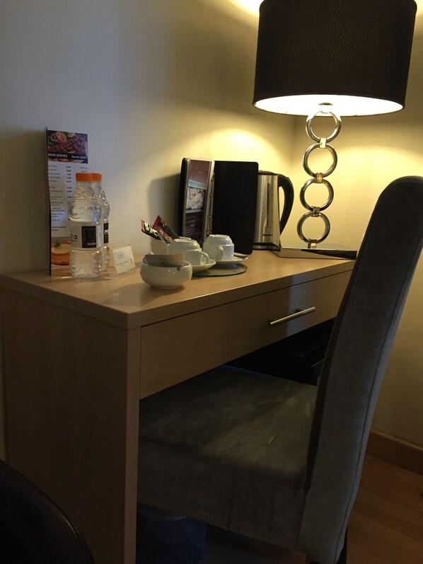 Mesa de quarto de hotel com uma luminária, uma garrafa de água, uma cafeteira e um quadro de cardápio.