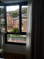 Quarto com uma grande janela que dá para uma área residencial com casas de tijolo e uma colina ao fundo.