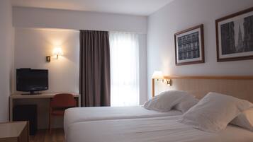 Quarto de hotel com duas camas, uma mesa de trabalho com televisão, uma cadeira e duas luminárias.