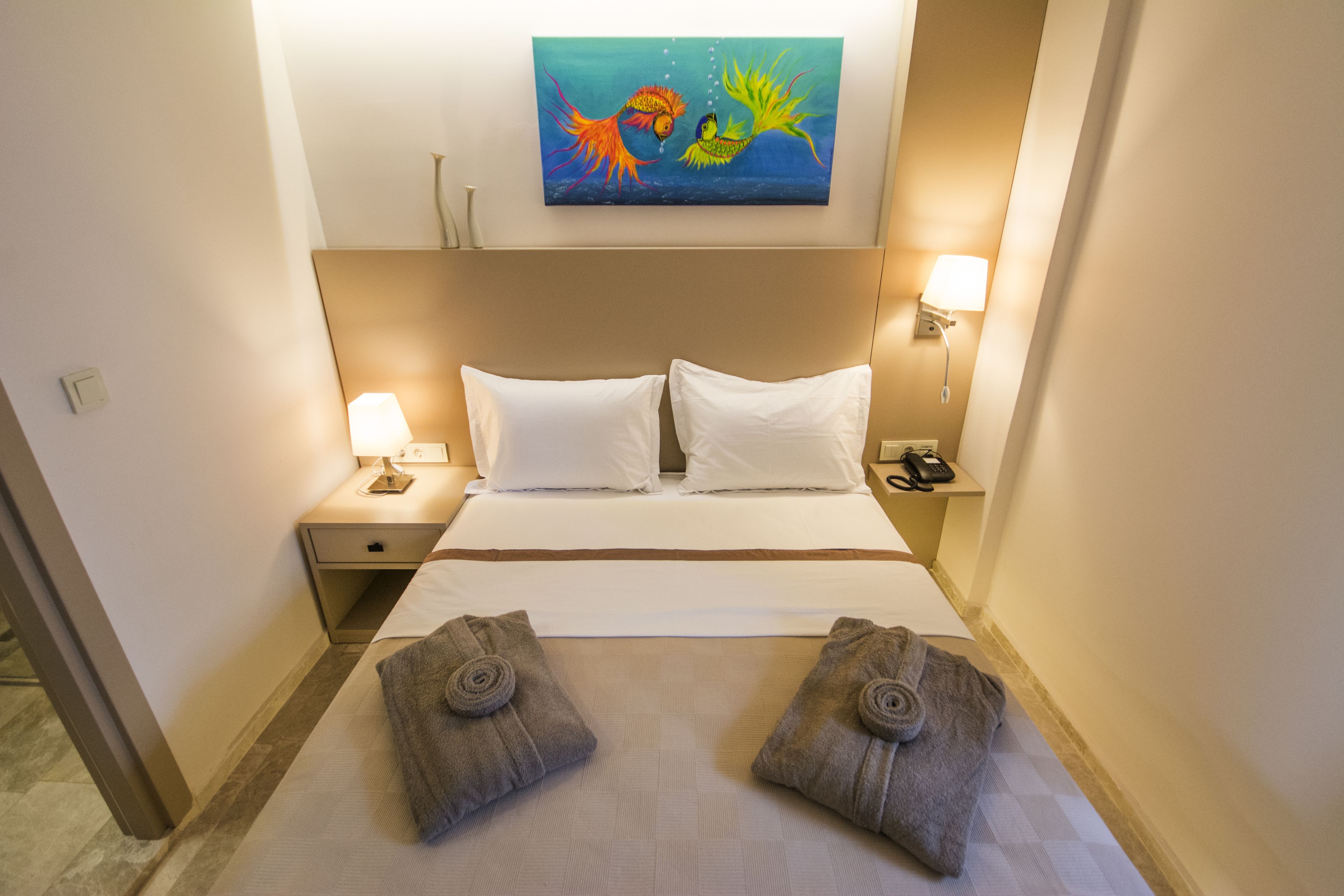 Quarto de hotel com uma cama, duas almofadas, mesa de cabeceira com abajur, um quadro sobre a cama e duas toalhas na cama.