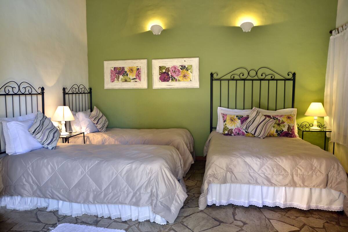 Quarto com duas camas, cada uma com cabeceira, mesa de cabeceira com luminária, e dois quadros florais emoldurados na parede.