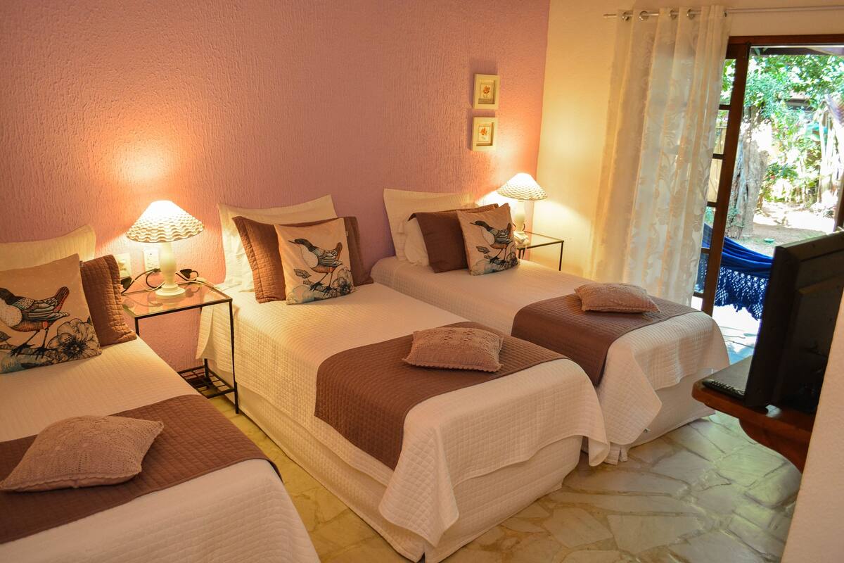 Quarto de hotel com duas camas, cada uma com travesseiro e luminária. Há uma televisão sobre um suporte de madeira e uma janela com vista para o verde lá fora.