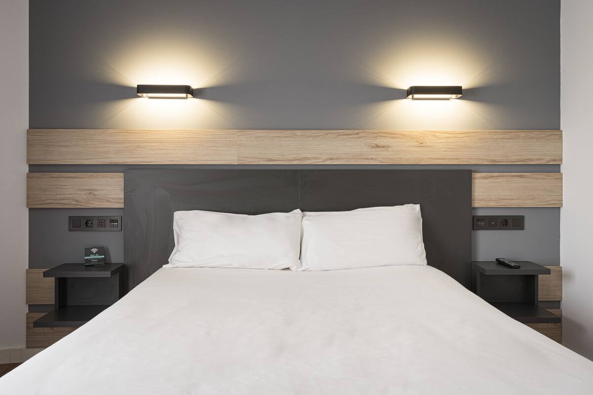 Quarto de hotel moderno com uma cama grande, duas mesas de cabeceira e um painel de madeira na cabeceira.