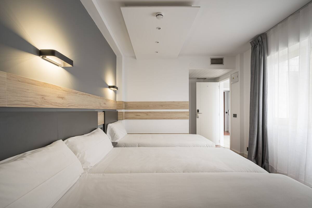 Quarto de hotel com duas camas, cabeceira de madeira, luminária e vista para um corredor.