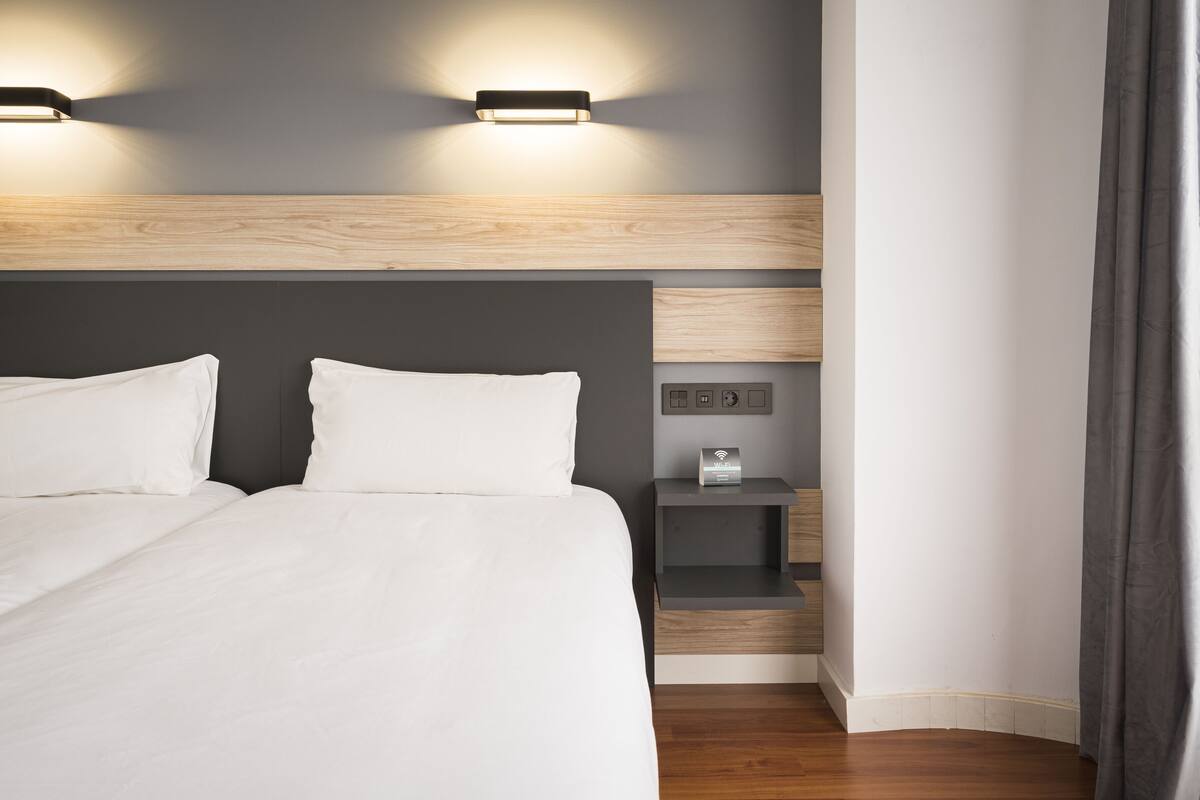 Quarto de hotel moderno com uma cama grande, uma mesa de cabeceira com abajur e um painel de controle.