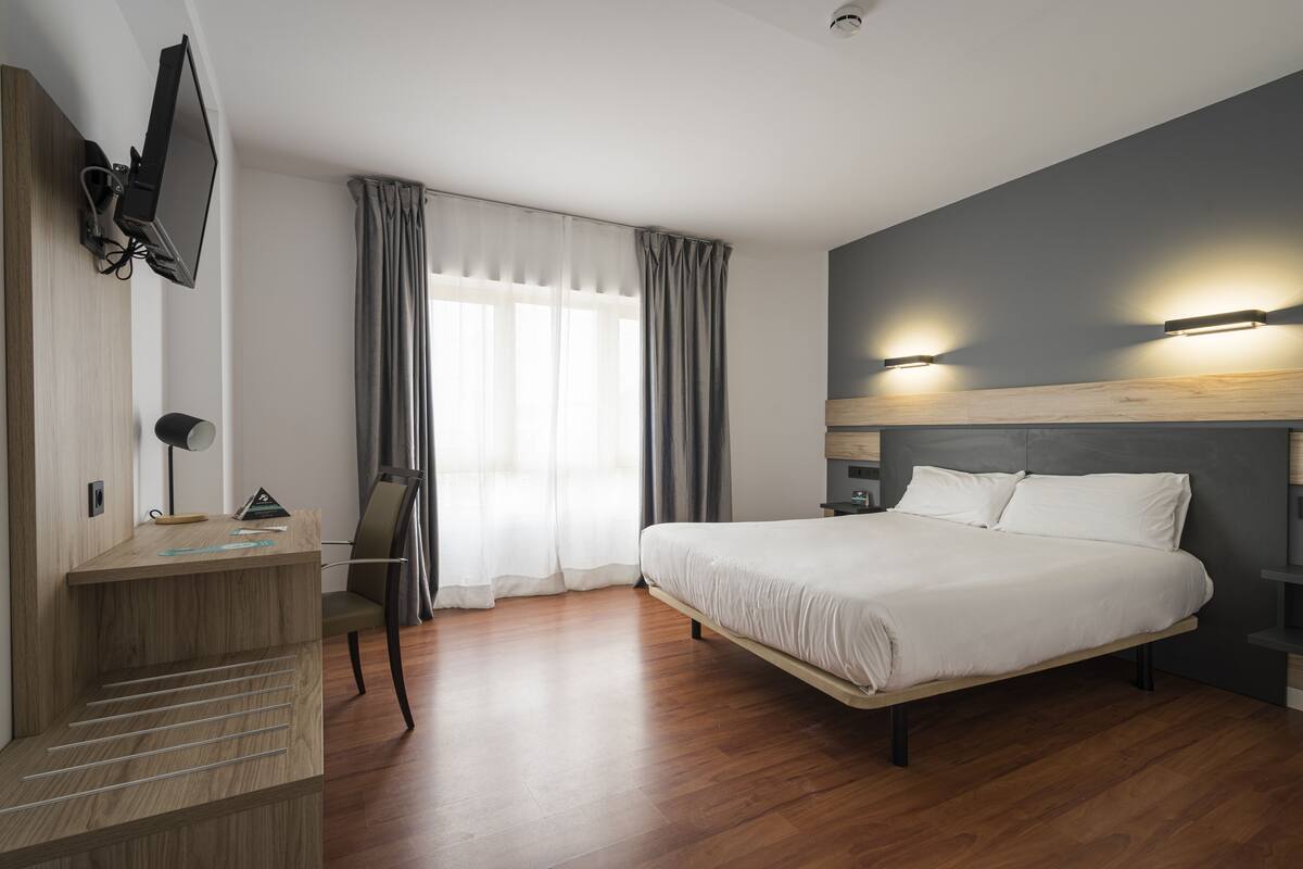 Quarto de hotel moderno com uma cama, uma escrivaninha com cadeira, uma televisão na parede e uma janela com cortinas.