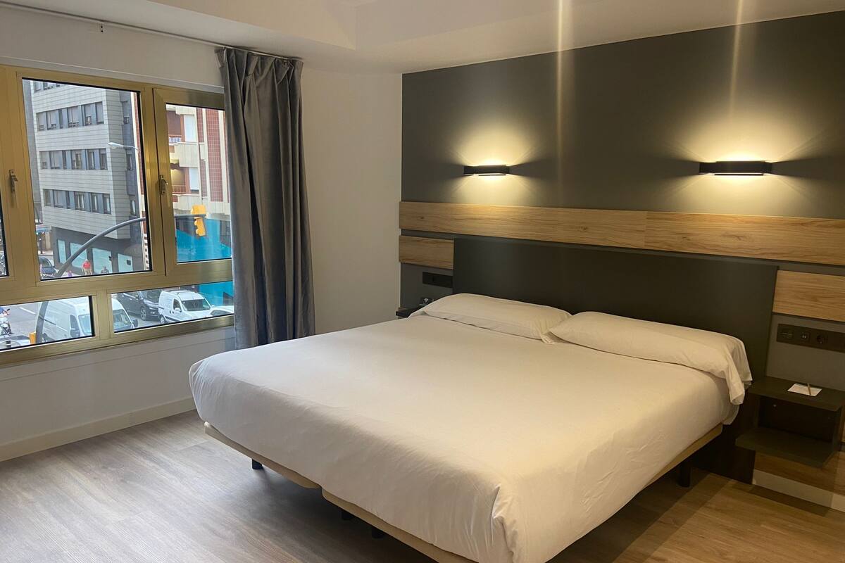 Quarto de hotel moderno com uma cama grande, duas luzes fixadas na parede, uma janela com vista para a rua e uma cabeceira de madeira.