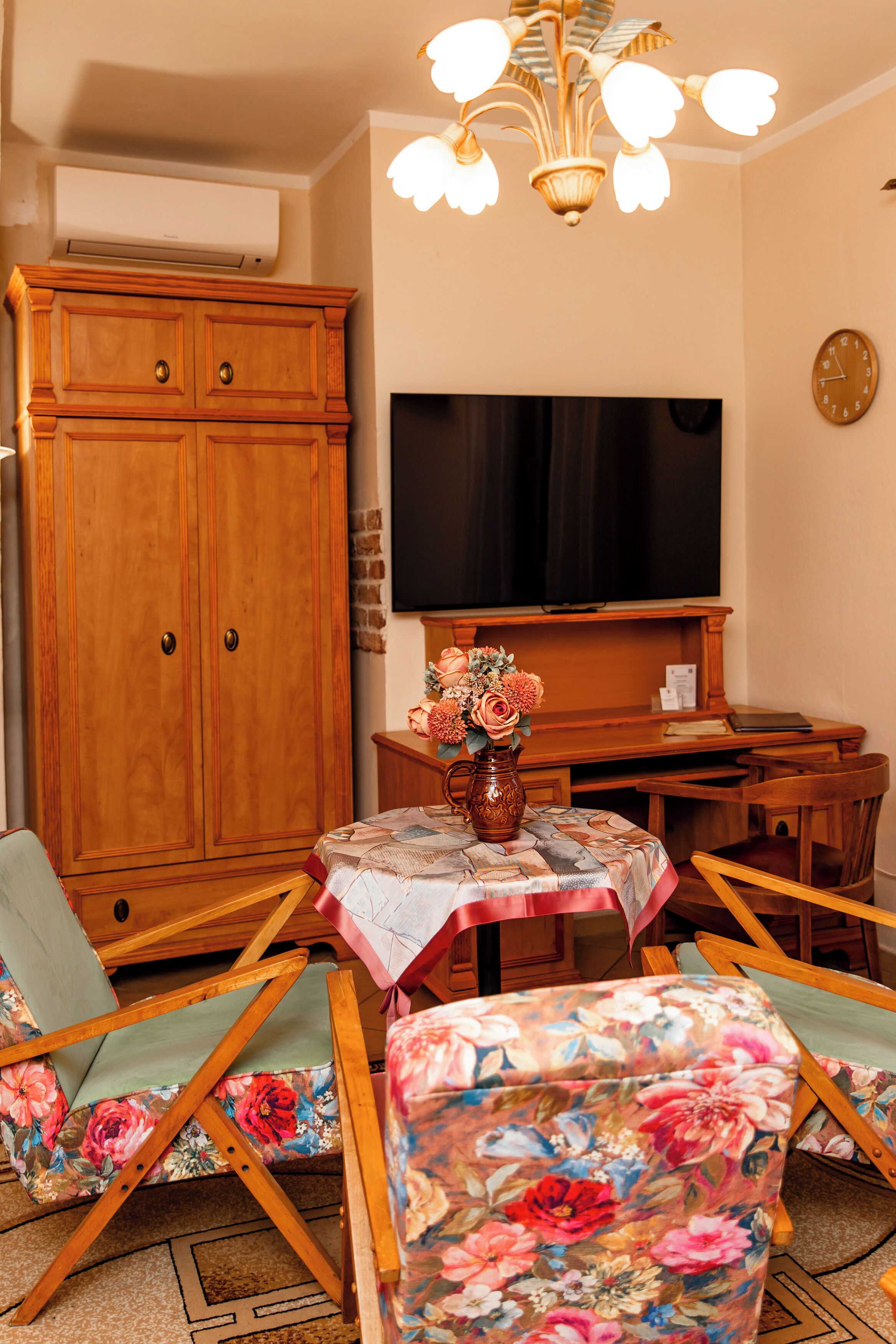 Uma sala de estar com televisão, armário de madeira, poltronas florais e uma mesa com um vaso.