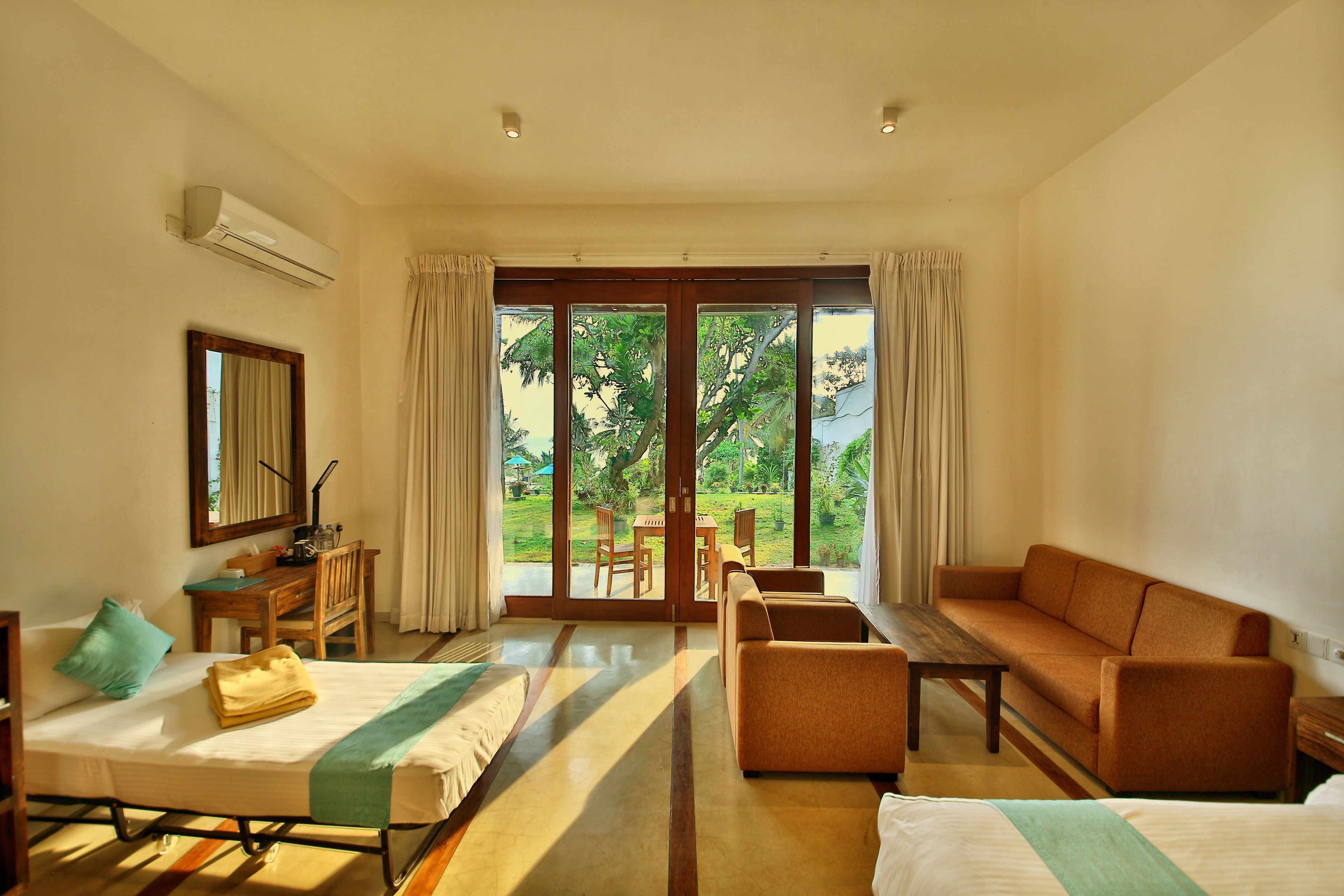 Quarto de hotel com cama, sofá, escrivaninha e vista para um jardim através de grandes janelas.