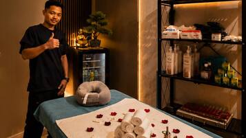 Uma mesa de massagem com toalha e pétalas de rosa, uma cadeira de massagem e uma prateleira com frascos e uma vela.