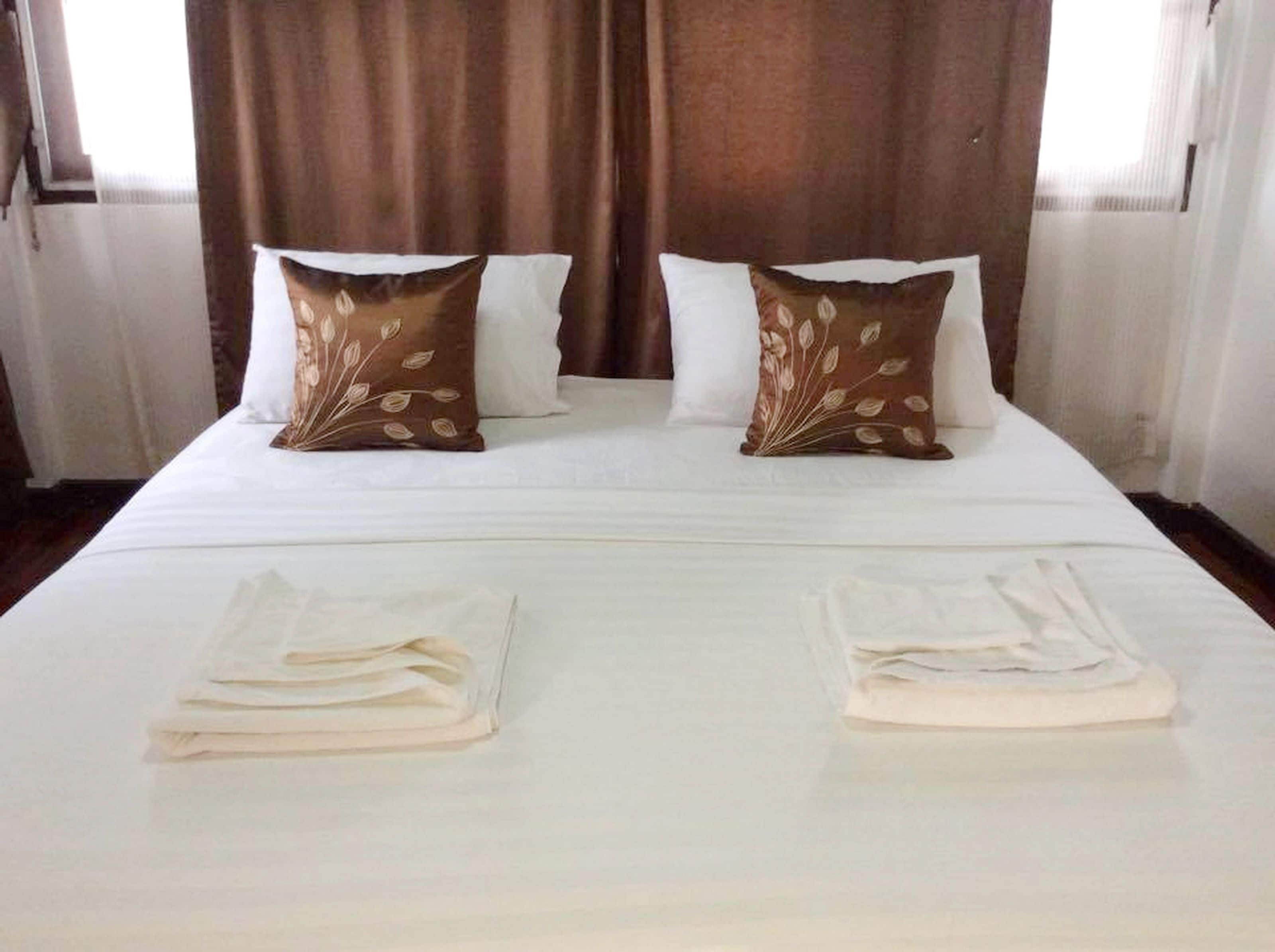Uma cama bem arrumada com duas almofadas, uma toalha dobrada e uma janela com cortinas.
