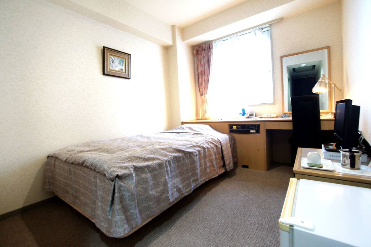 Quarto de hotel pequeno com uma cama de solteiro, uma escrivaninha com computador, uma janela com cortinas e uma mesinha de cabeceira.