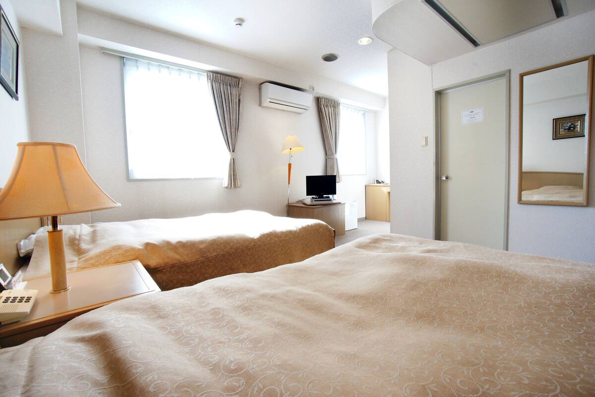 Quarto de hotel com duas camas, uma escrivaninha com computador, uma luminária e uma janela com cortinas.
