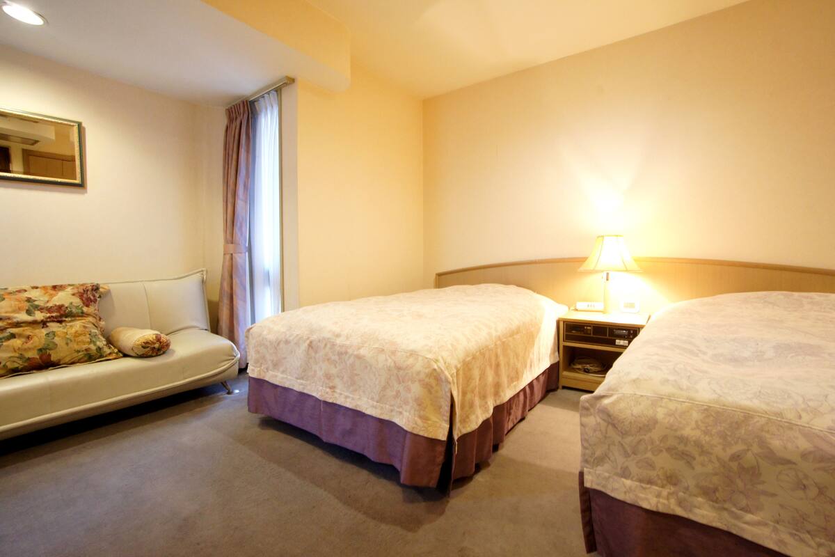Quarto de hotel com duas camas, um sofá, uma mesa de cabeceira e uma luminária.