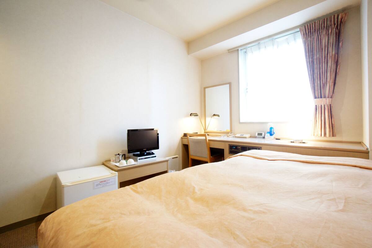 Quarto de hotel com cama, escrivaninha com computador, televisão e janela com cortinas.