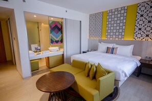 Quarto de hotel com cama, sofá, mesinha, banheiro com pia e espelho, e parede estampada.