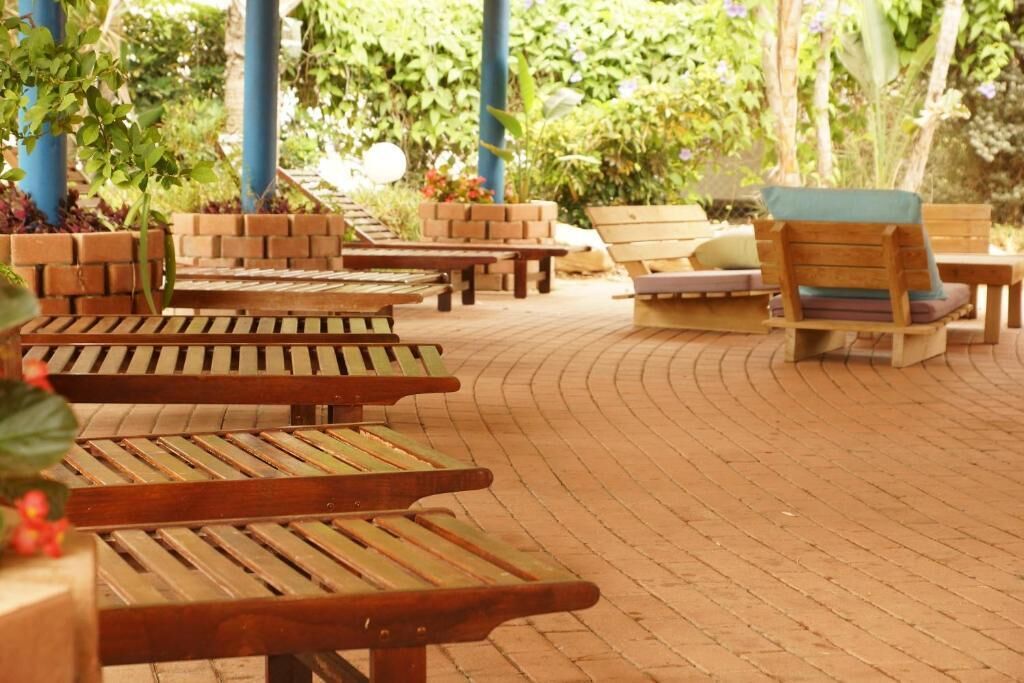 Espaço ao ar livre com bancos e mesas de madeira, cercado por vegetação e vasos de barro.