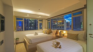 Quarto de hotel com duas camas, mesa de cabeceira, luminária e vista da cidade iluminada à noite.
