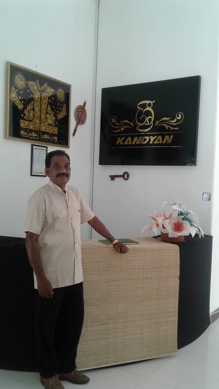 Um homem em frente a um balcão de recepção, com uma obra de arte emoldurada acima e uma placa com o nome 'KANDYAN' na parede.