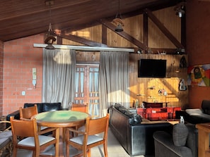 Uma sala de estar com teto de madeira, uma TV fixada na parede, um sofá, uma mesa de jantar com cadeiras e uma janela com cortinas.