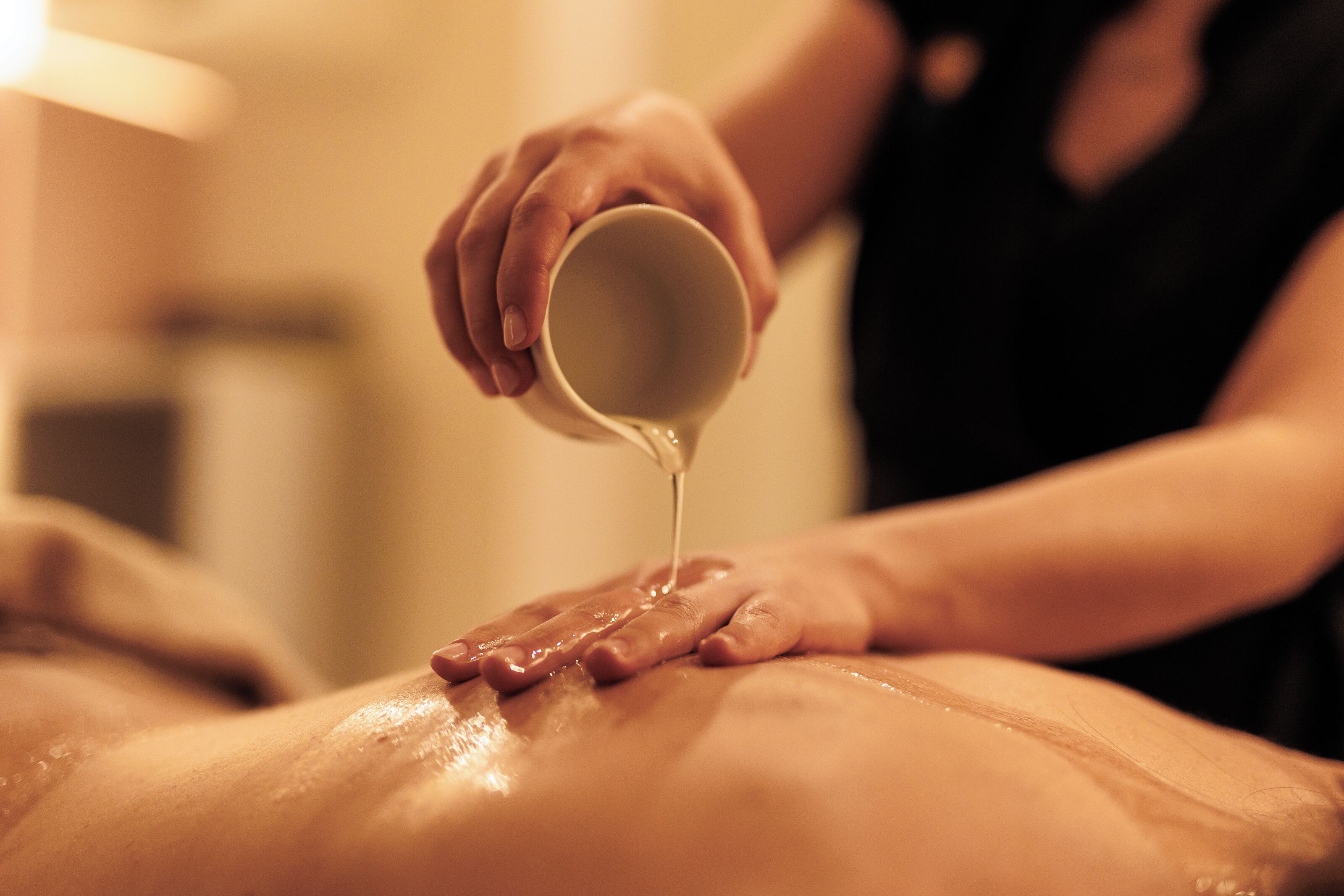 Close-up de mãos recebendo massagem, com líquido sendo derramado nas costas.